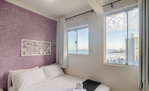 Quarto com cama, janela com vista para edifícios e mar, um objeto decorativo na parede e uma toalha sobre a cama.