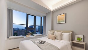 Quarto de hotel moderno com uma cama grande, uma mesa de cabeceira, uma luminária e uma janela com vista para a cidade, além de um quadro na parede.
