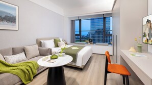 Quarto de hotel moderno com cama, sofá, escrivaninha e uma grande janela com vista para a cidade.