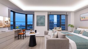 Quarto de hotel moderno com uma cama grande, um sofá, uma mesa de escritório e um espaço de jantar. O quarto possui janelas amplas com vista para a cidade.