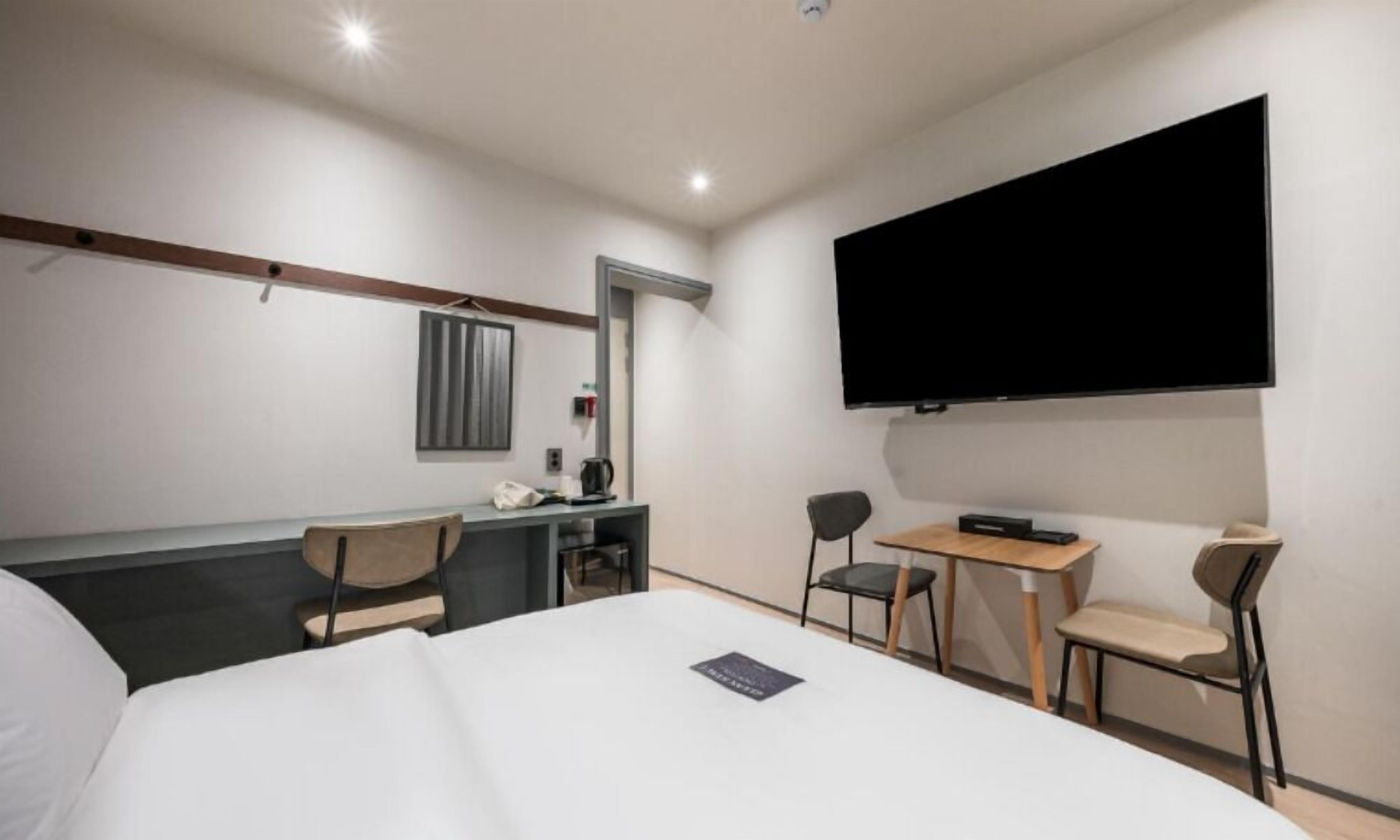 Quarto de hotel com cama, escrivaninha, duas cadeiras, televisão e espelho.