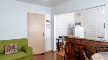 Um espaço de estar compacto com sofá verde, pequena área de cozinha com micro-ondas e frigorífico, e mesa de jantar de madeira.