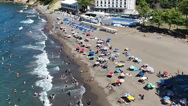 Uma praia com muitos guarda-sóis e pessoas, um edifício com piscina e uma encosta arborizada.