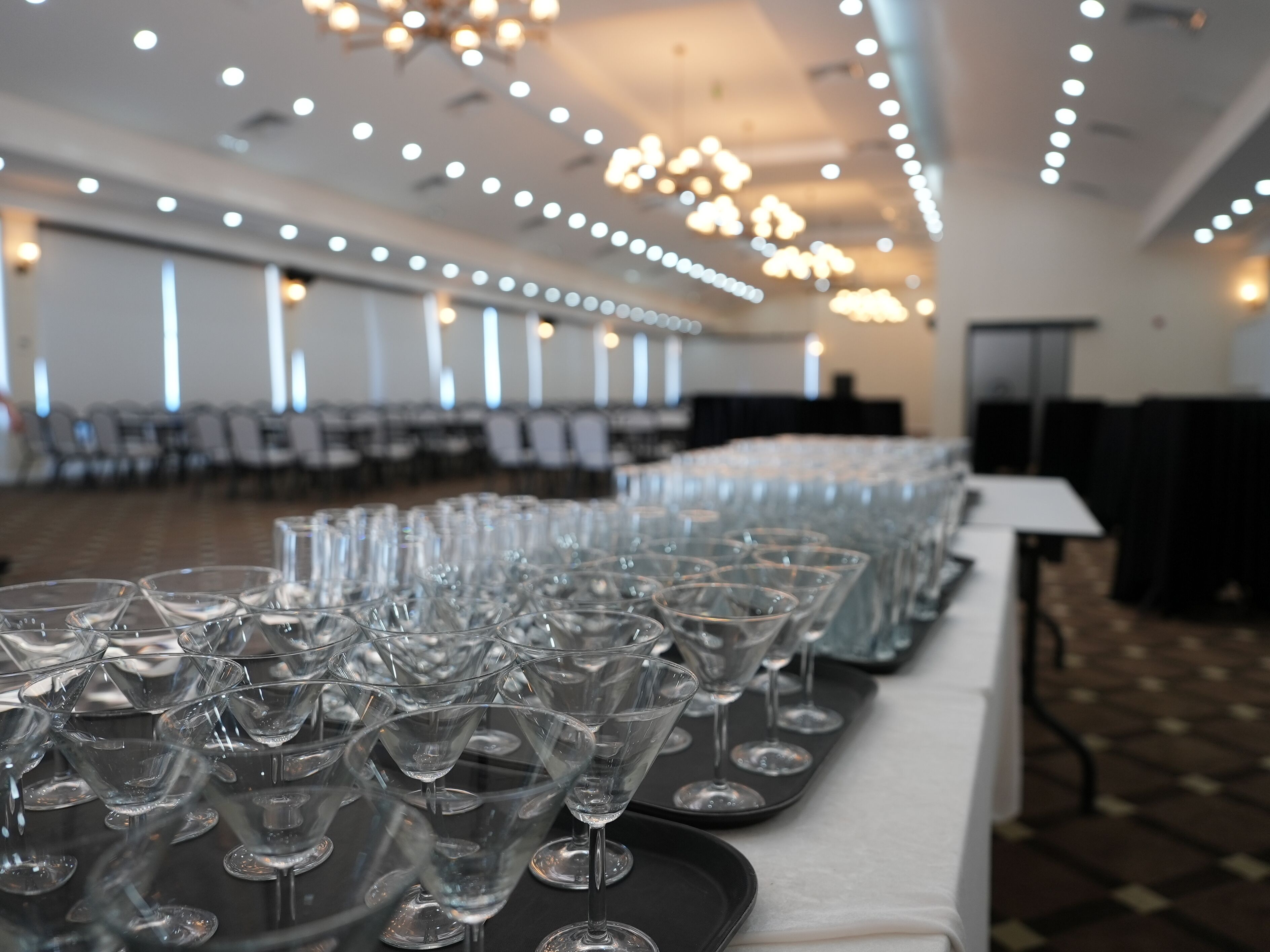 Uma sala de banquetes com fileiras de copos de vinho vazios em bandejas, uma mesa comprida e um teto com iluminação embutida.