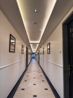 Corredor comprido e estreito de um hotel, com piso de azulejos, quadros nas paredes e iluminação embutida.