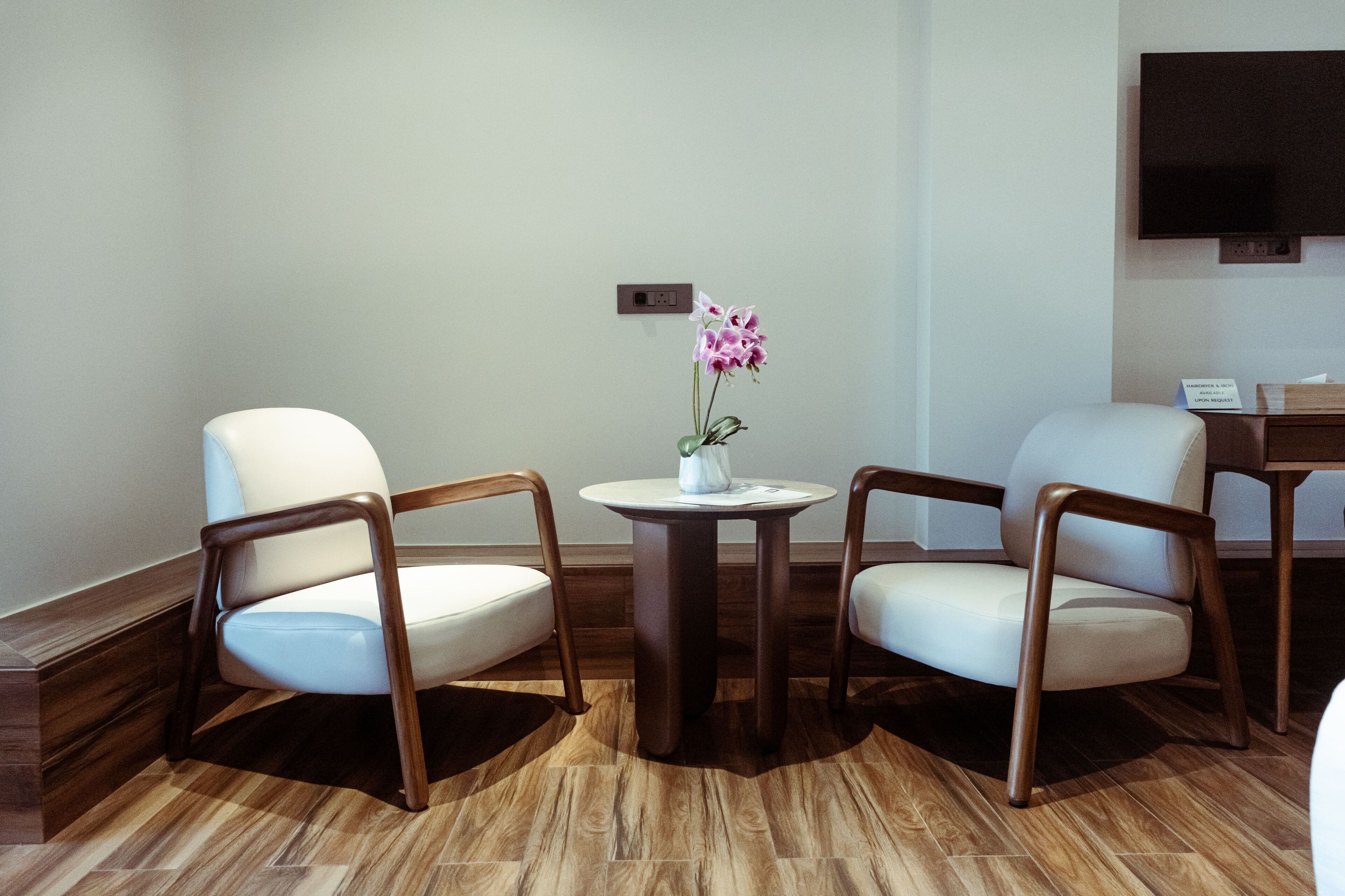 Quarto com duas cadeiras brancas, uma mesa redonda pequena com um vaso de flores e uma TV de tela plana na parede.