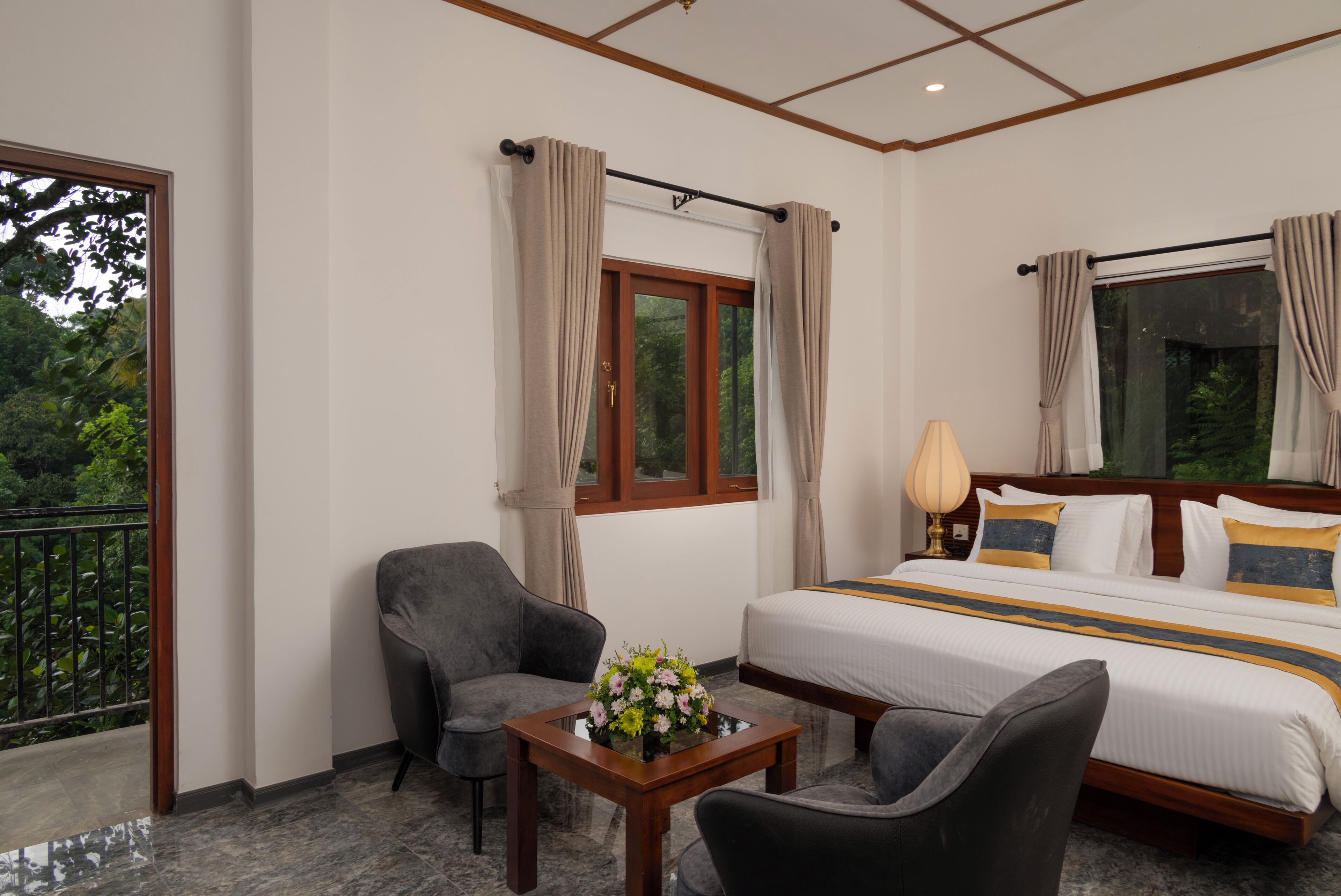 Quarto de hotel com cama, duas cadeiras, uma mesa com arranjo floral, janela com cortinas e varanda com vista para o verde.