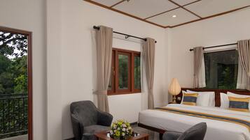 Quarto de hotel com cama, duas cadeiras, uma mesa com arranjo floral, janela com cortinas e varanda com vista para o verde.
