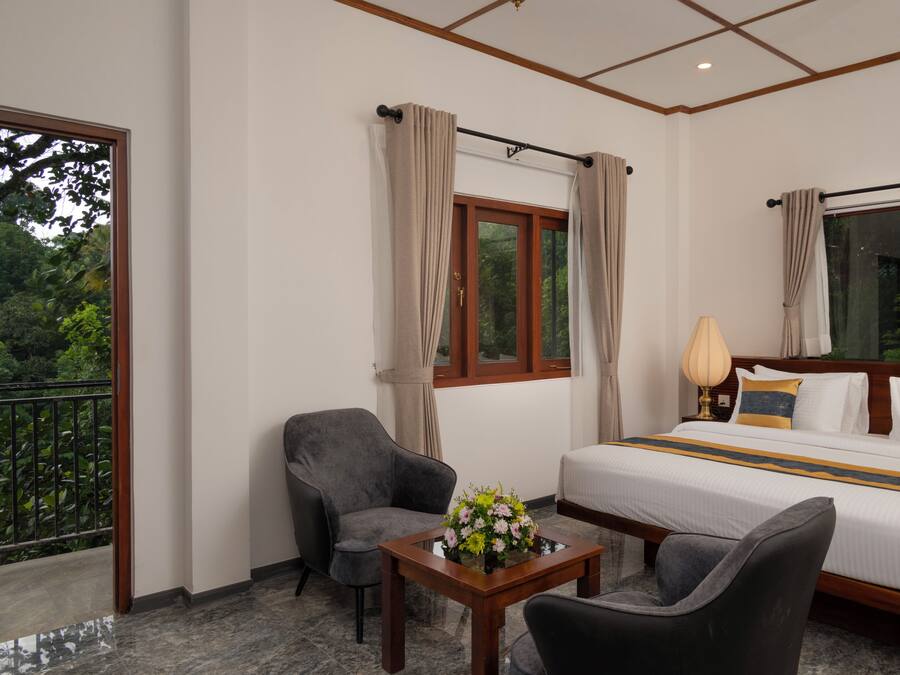 Quarto de hotel com cama, duas cadeiras, uma mesa com arranjo floral, janela com cortinas e varanda com vista para o verde.