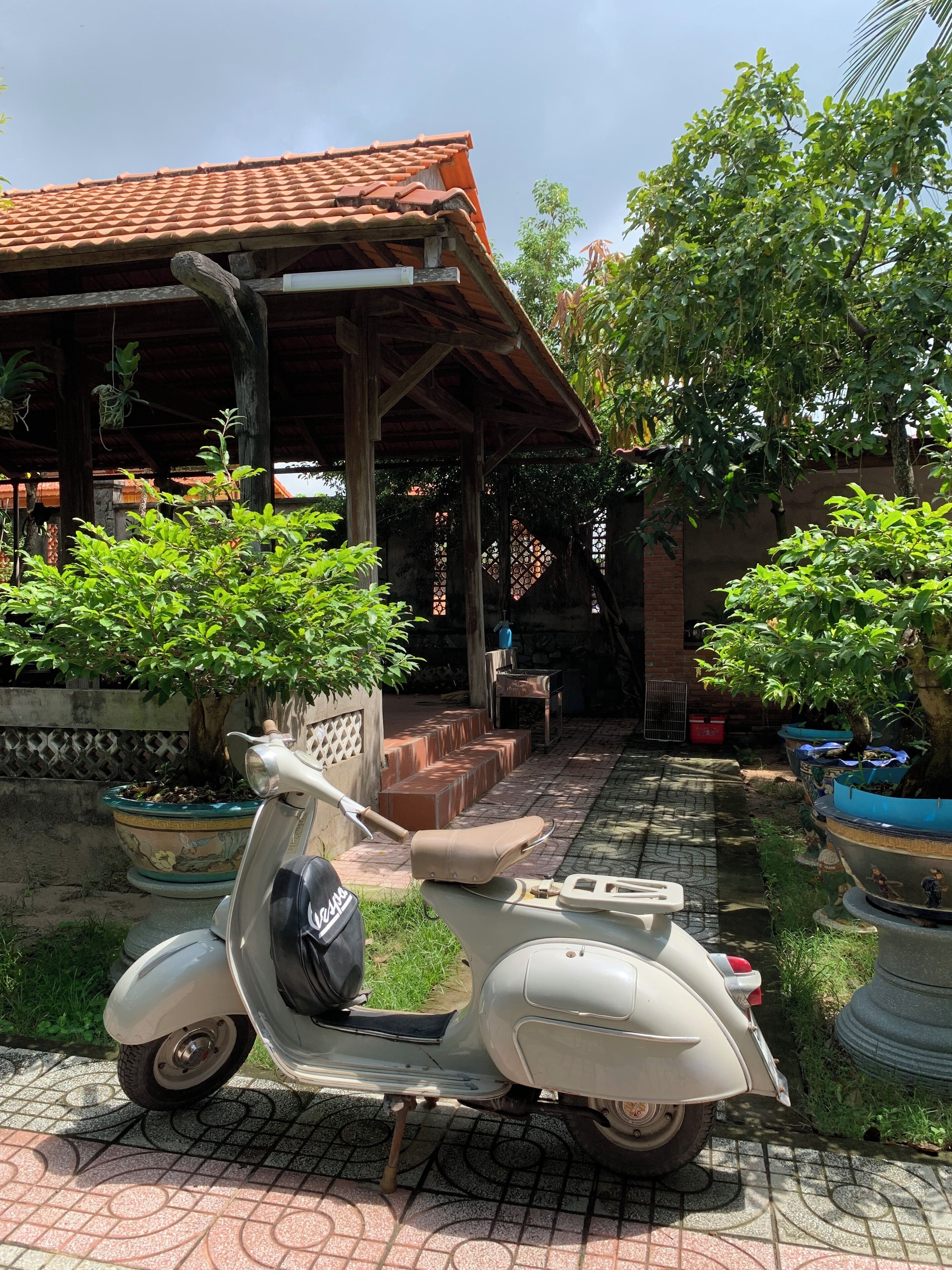 Uma scooter branca estacionada em um pátio com piso de azulejos, cercada por plantas em vasos e um edifício com telhado de azulejos.