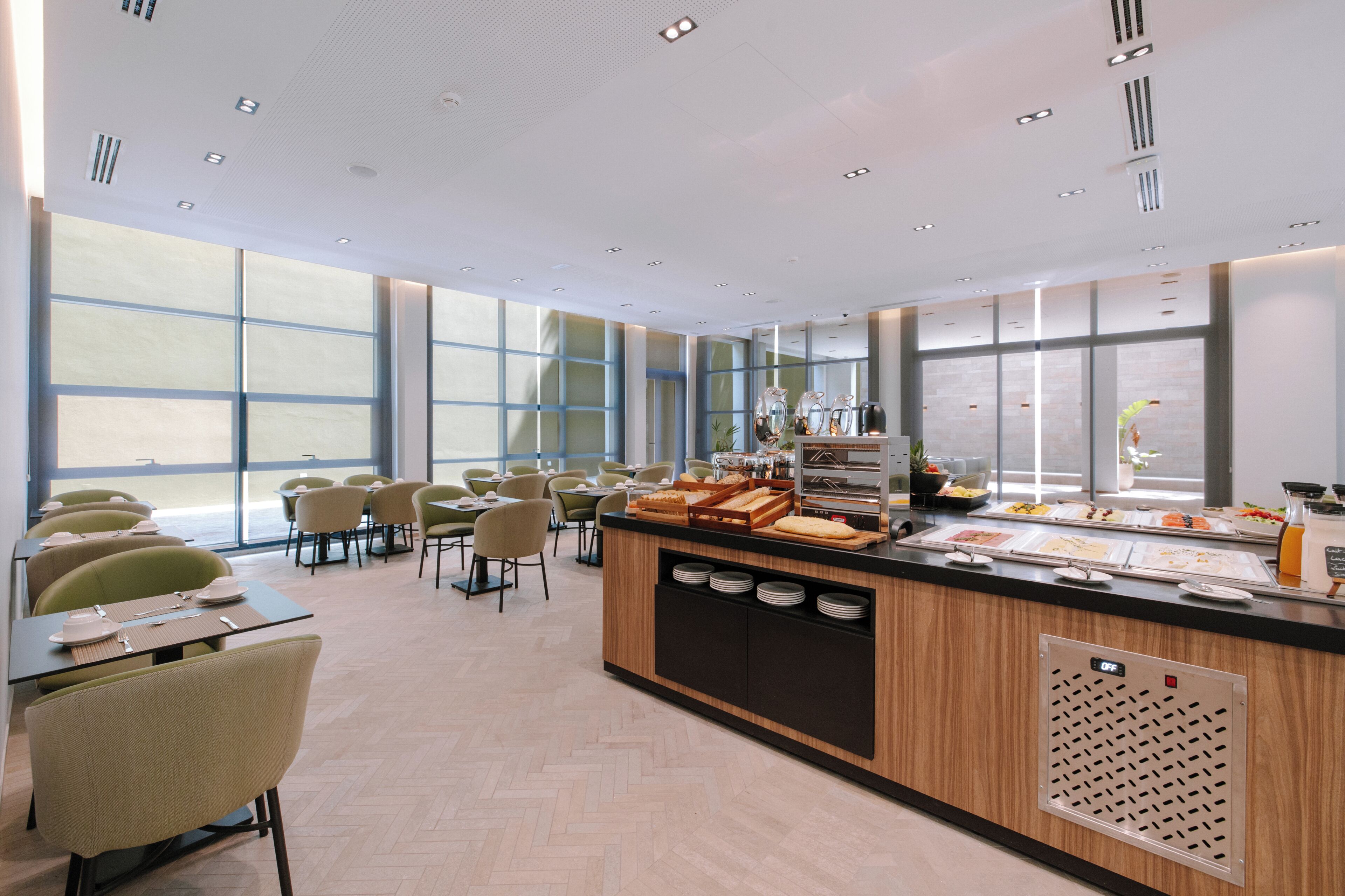 Uma área de refeições moderna em hotel, com um balcão central que serve café da manhã, rodeada por mesas e cadeiras.