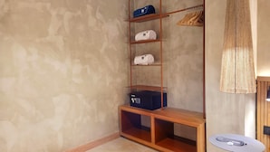 Canto de um quarto com uma estante de madeira encostada em uma parede texturizada, um candeeiro tecido e uma pequena mesa redonda com superfície preta.