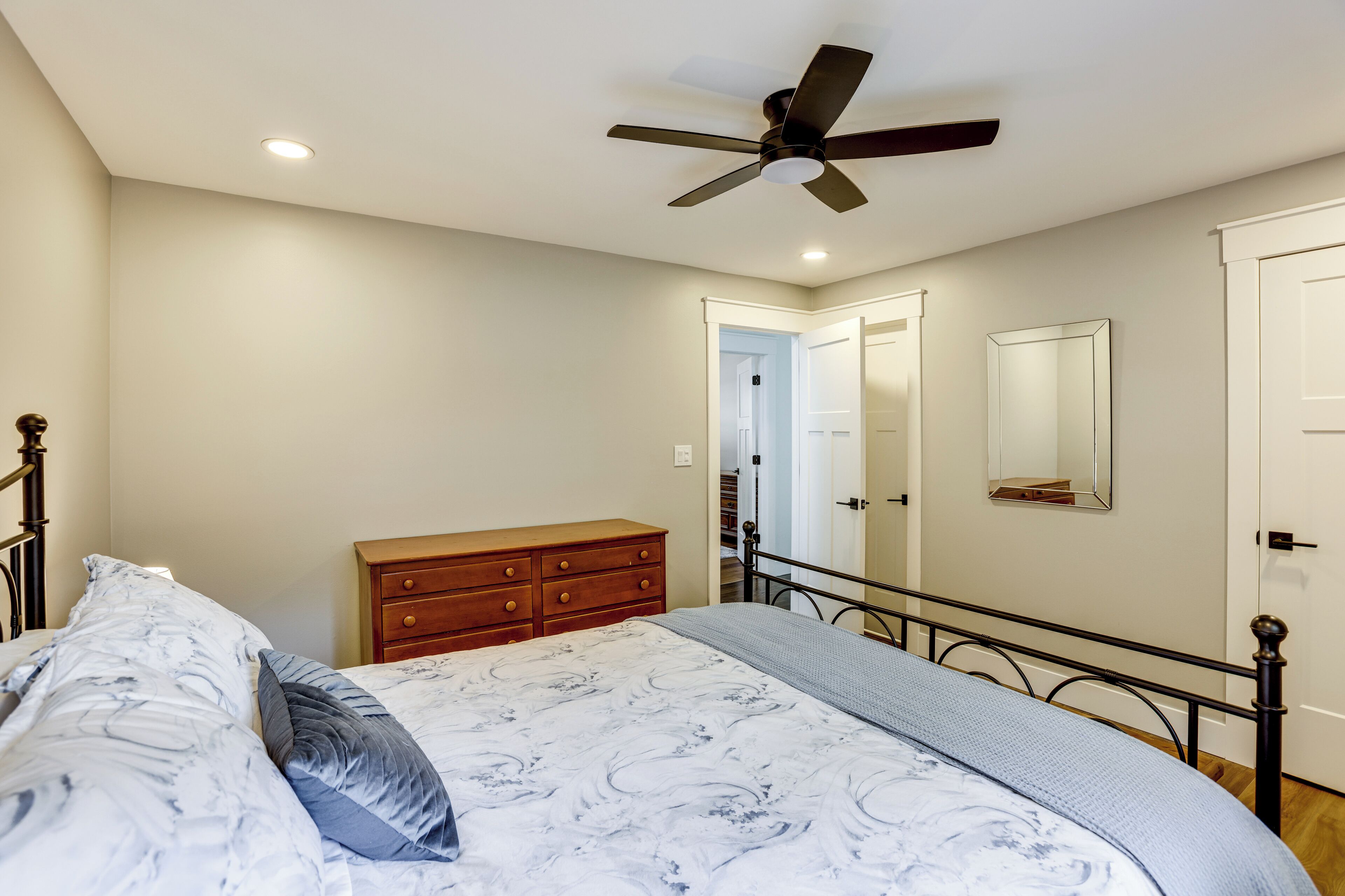 Quarto com cama, penteadeira de madeira, ventoinha no teto e porta que dá para outro cômodo.