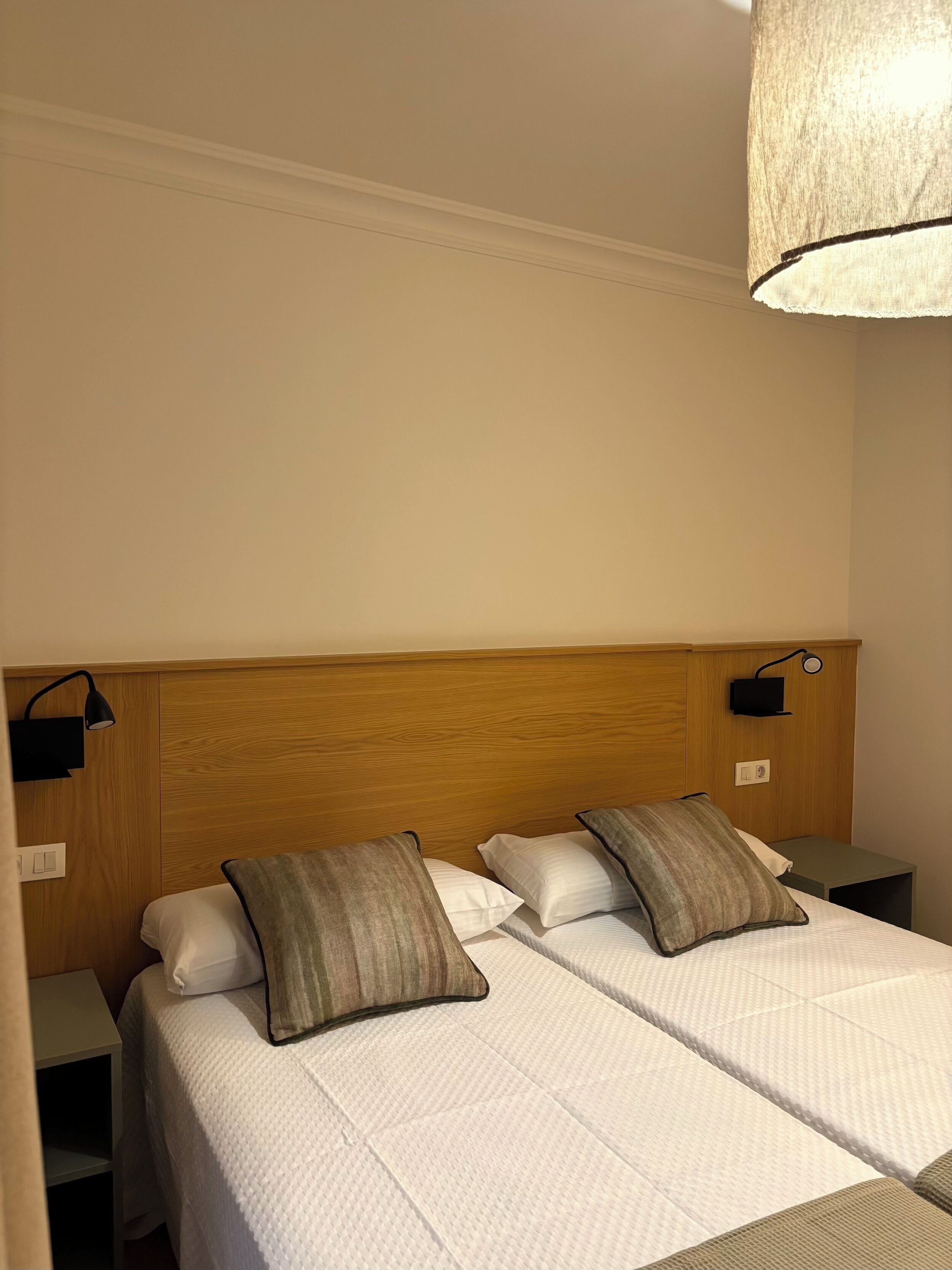 Quarto de hotel com cabeceira de madeira, duas camas com roupa de cama branca e uma luminária de cabeceira.
