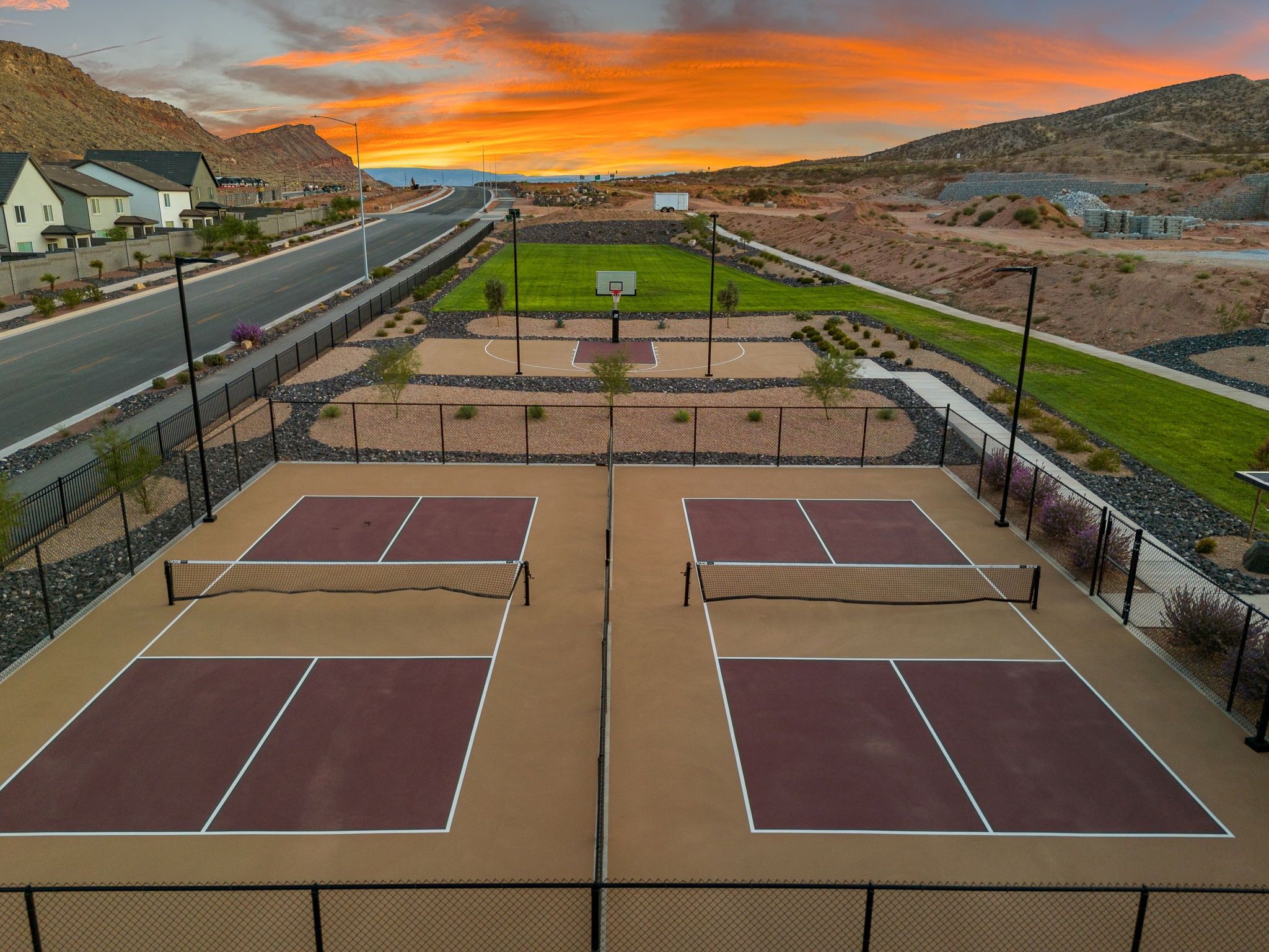 Quadra de basquete ao ar livre com duas quadras, cercada por uma grade, com uma estrada e casas ao fundo.