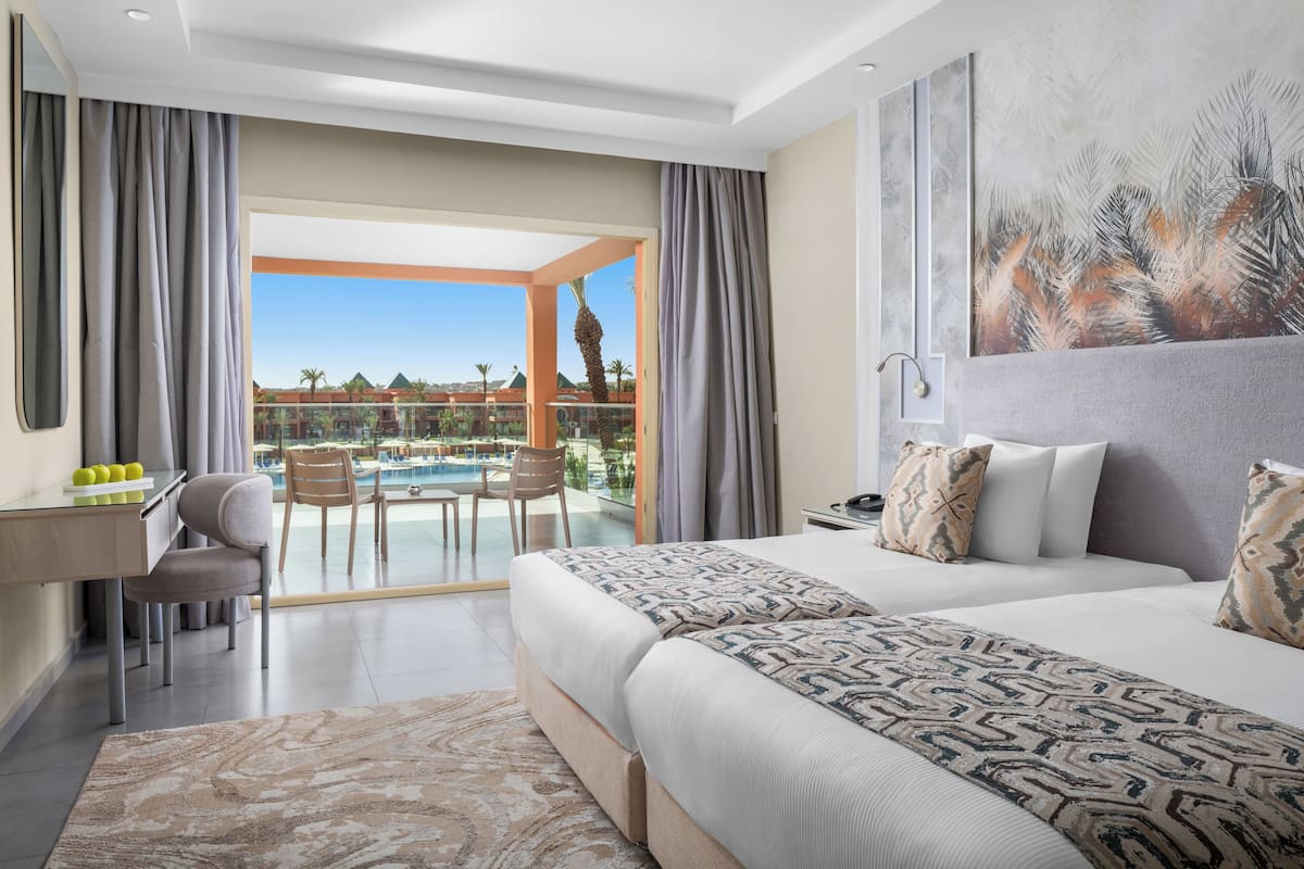 Quarto de hotel com uma cama grande, uma mesa com cadeira e vista para uma piscina e palmeiras.