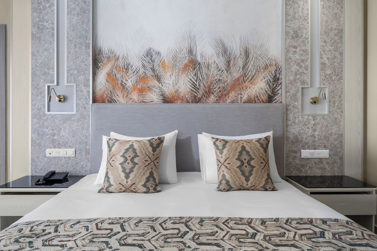 Quarto de hotel moderno com uma cama grande, duas almofadas estampadas iguais, uma mesa de cabeceira com telefone e luminária fixa na parede.