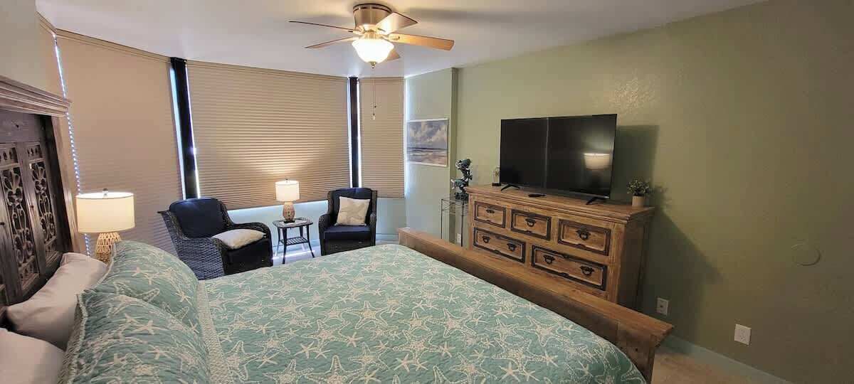 Quarto com cama, televisão sobre uma cômoda, cadeira, abajur e janela com persianas.