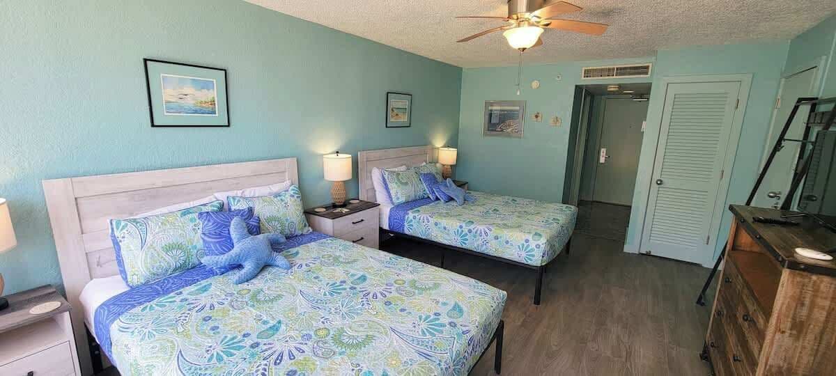 Quarto com duas camas, cada uma com mesa de cabeceira e luminária, um ventilador de teto e uma porta que dá para outro cômodo.