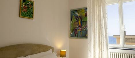 Quarto de hotel com cama, mesa de cabeceira com abajur, dois quadros na parede e janela com cortinas.