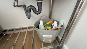 Um balde rotulado 'Produtos de Limpeza da Cozinha' sob uma pia com uma mangueira flexível.