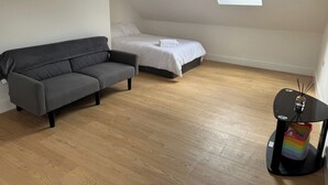 Quarto com sofá cinza, cama com roupa de cama branca, uma pequena mesa com um cesto e um claraboia.