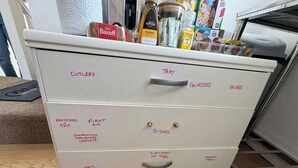 Baú branco com gavetas identificadas para talheres, bandeja, copos, canecas, kit de primeiros socorros, blocos de notas, folhetos informativos, louças, secador de cabelo, lençol plano para sofá-cama e tecido.