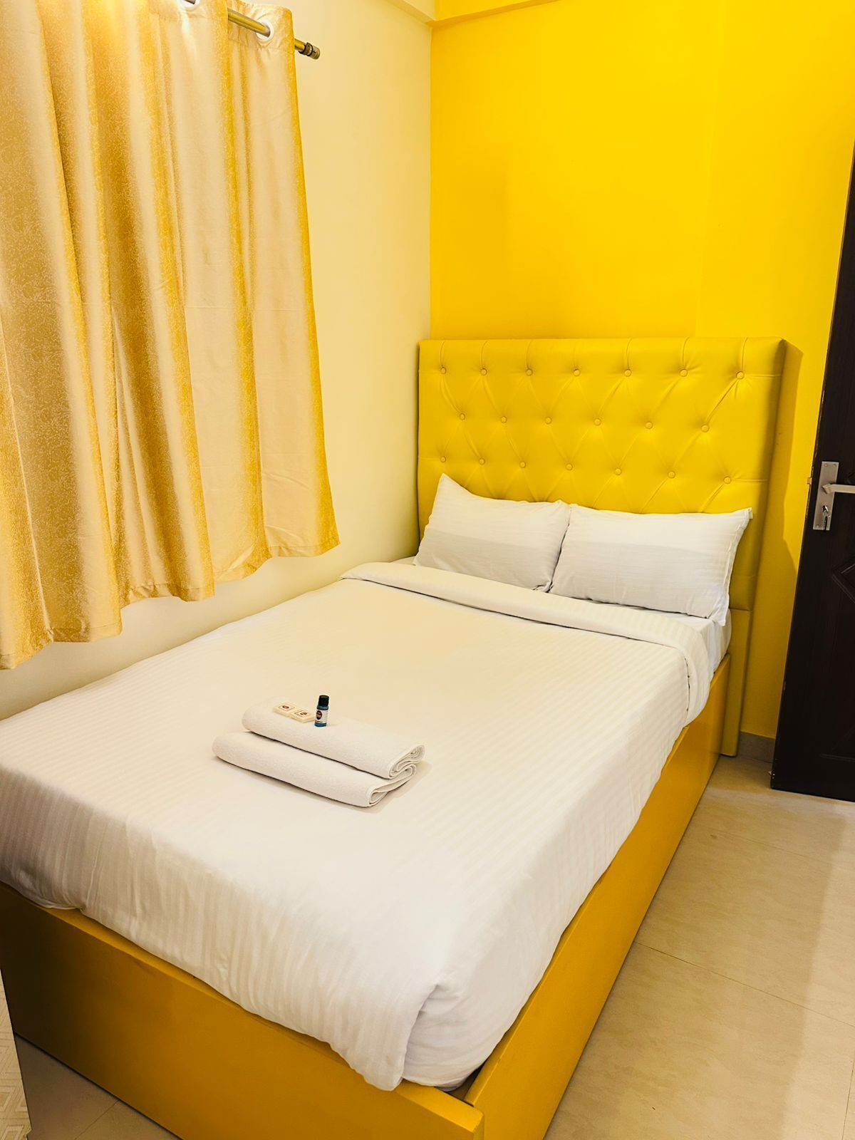 Uma cama de solteiro com cabeceira amarela, roupa de cama branca e uma toalha enrolada sobre a cama.