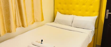 Uma cama de solteiro com cabeceira amarela, roupa de cama branca e uma toalha enrolada sobre a cama.
