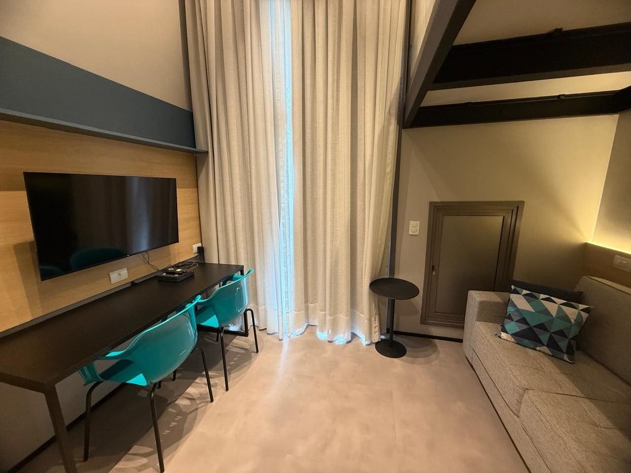 Uma sala de estar moderna com uma televisão de tela plana, poltronas verde-água, um sofá com almofadas estampadas e uma porta.