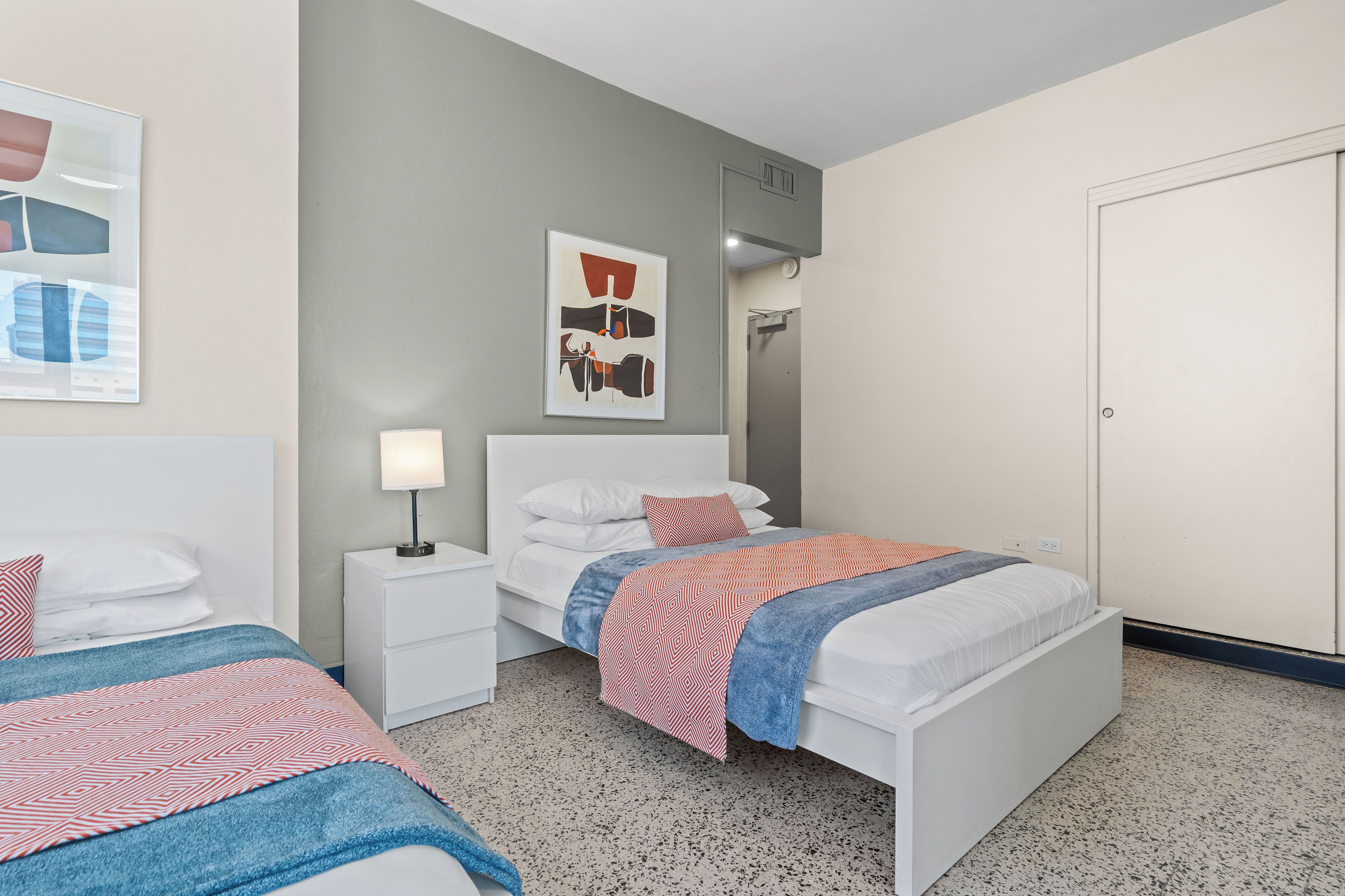 Quarto de hotel moderno com duas camas, uma mesa de cabeceira, uma luminária na parede e obras de arte.