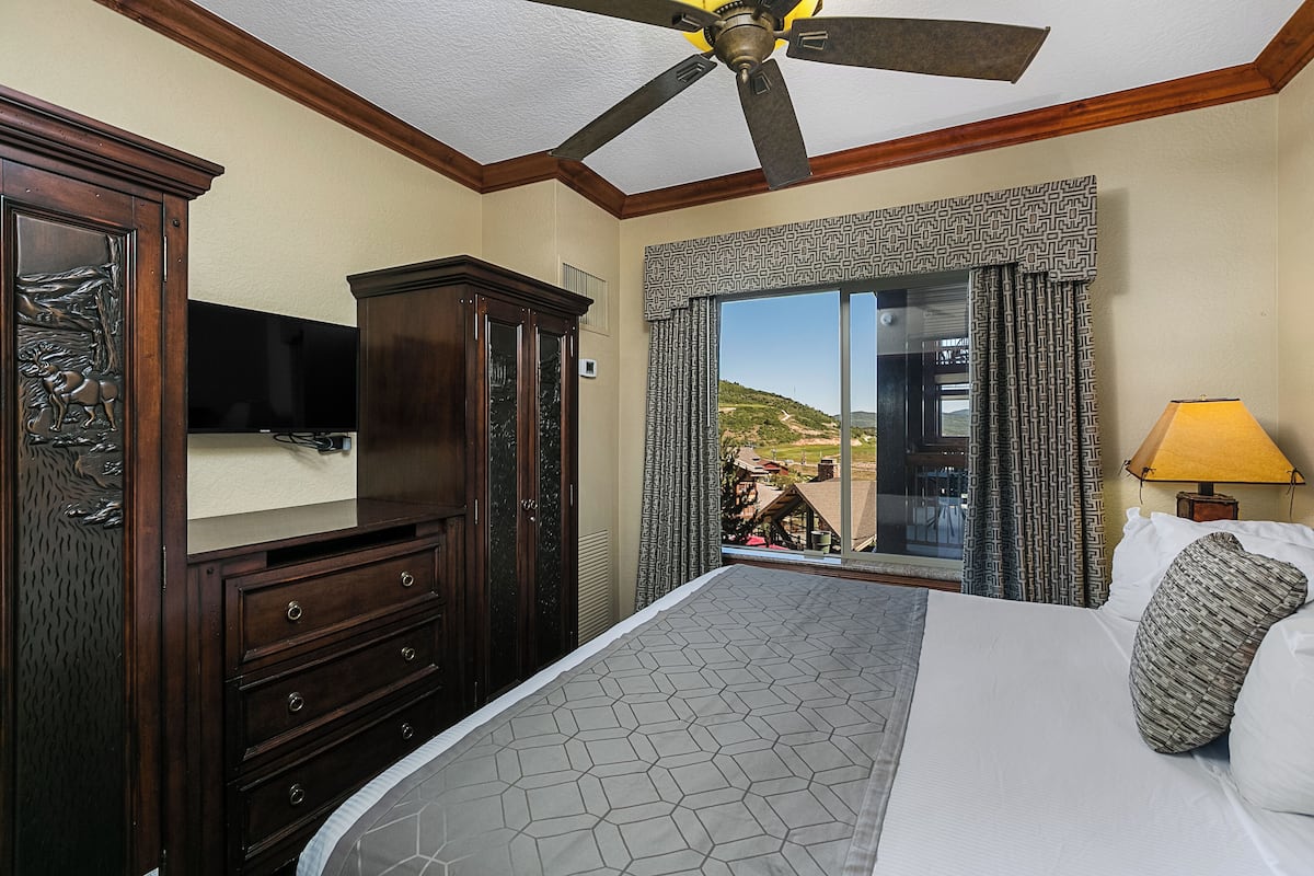 Quarto de hotel com uma cama grande, um ventilador de teto, uma TV, uma cômoda e uma janela com vista para uma colina.