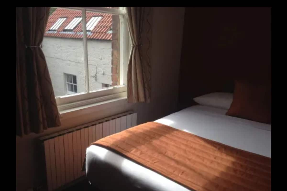 Quarto com uma cama, uma janela com cortinas e vista para um edifício com telhado vermelho.