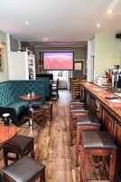 Área de bar com balcão, banquetas e uma televisão exibindo um evento esportivo.