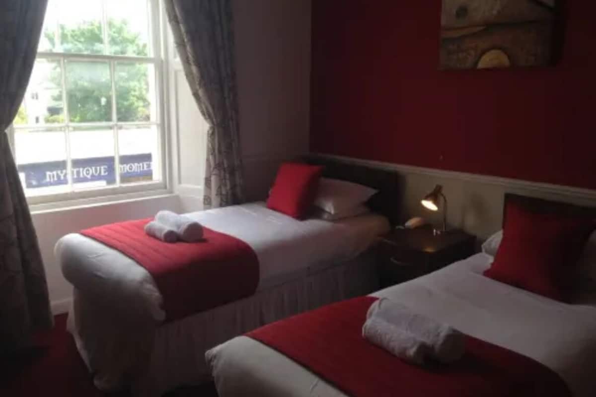 Quarto de hotel com duas camas, roupa de cama vermelha, uma janela com cortinas e uma mesa de cabeceira com abajur.