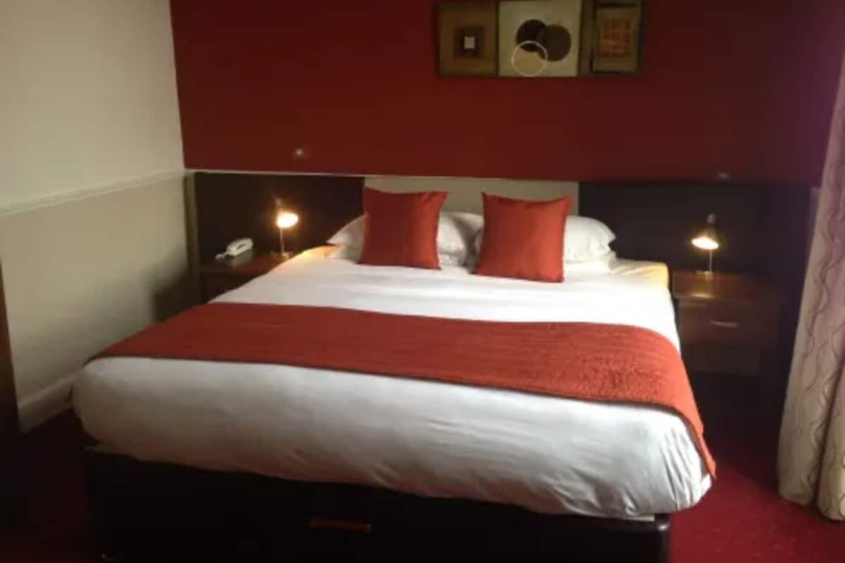 Uma cama bem arrumada com um cobertor vermelho e duas almofadas, uma mesinha de cabeceira com uma luminária, e uma parede com quadros.