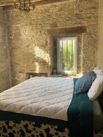 Um quarto com parede de pedra, janela com grades, cama com colcha branca, travesseiro azul e manta verde.
