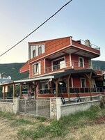 Um edifício de dois andares com varanda e área coberta, rodeado por vegetação e colinas.
