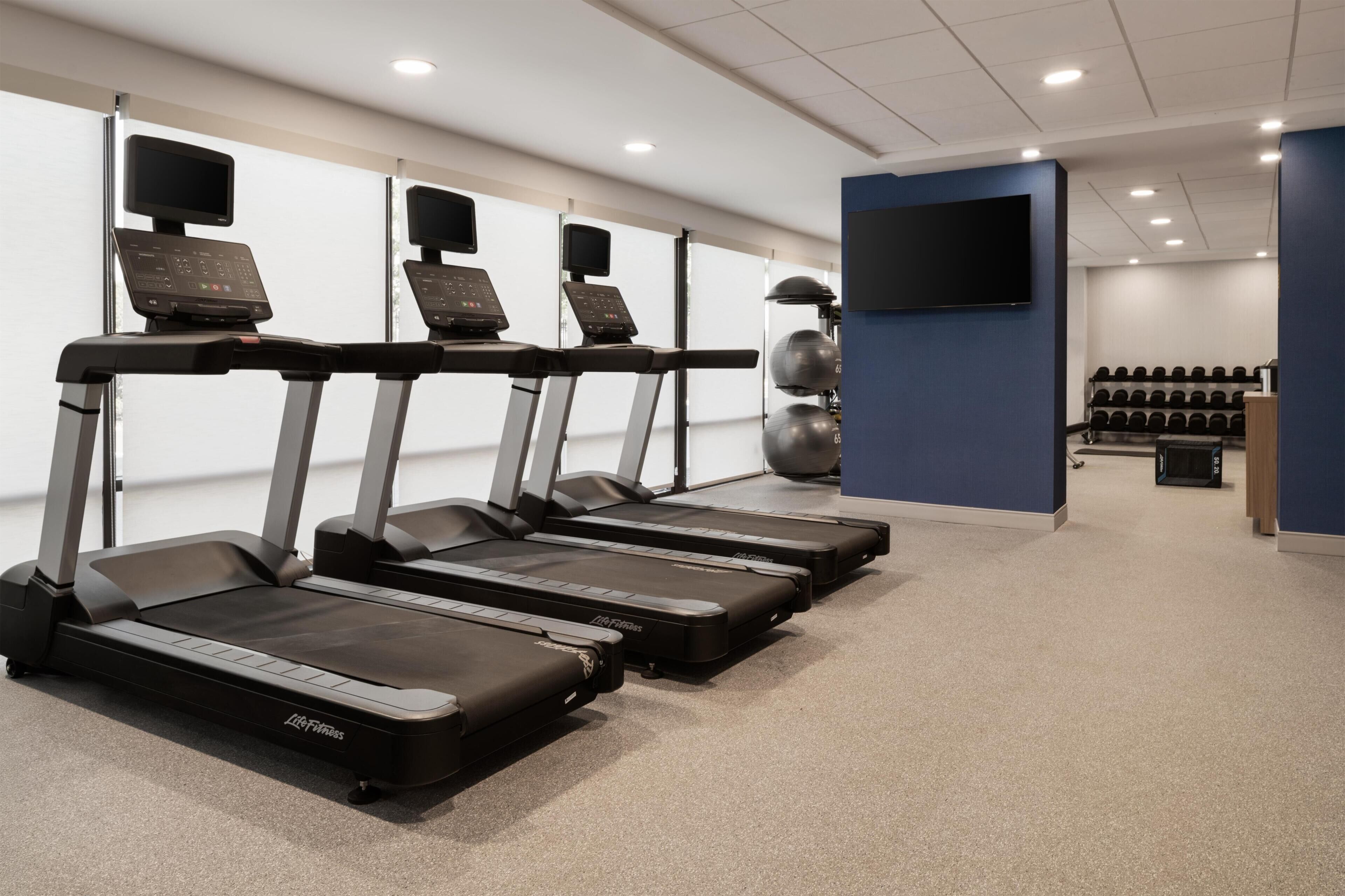 Centro de fitness com esteiras, área de musculação e tela de televisão.