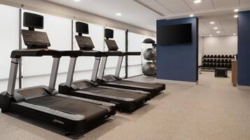 Centro de fitness com esteiras, área de musculação e tela de televisão.