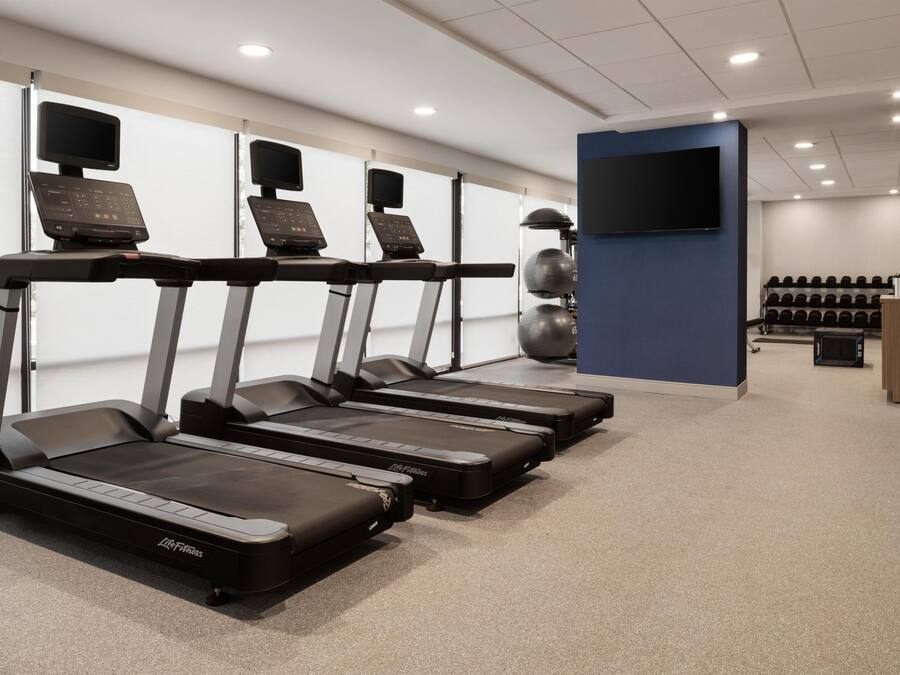 Centro de fitness com esteiras, área de musculação e tela de televisão.