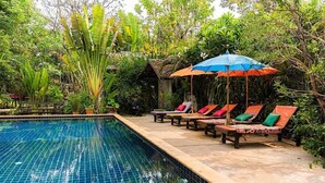 Piscina com piso em azulejo, rodeada por espreguiçadeiras com almofadas coloridas e um guarda-sol azul para dar sombra.