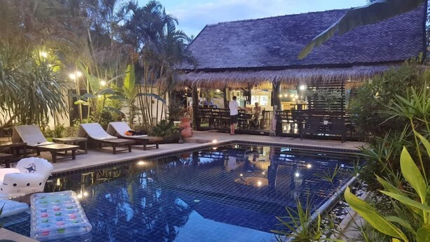 Uma área de piscina com espreguiçadeiras, bar e um edifício com telhado de palha.