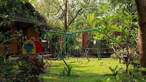 Um parquinho com balanços e um brinquedo de escalada em uma área ajardinada, com um edifício de madeira ao fundo.