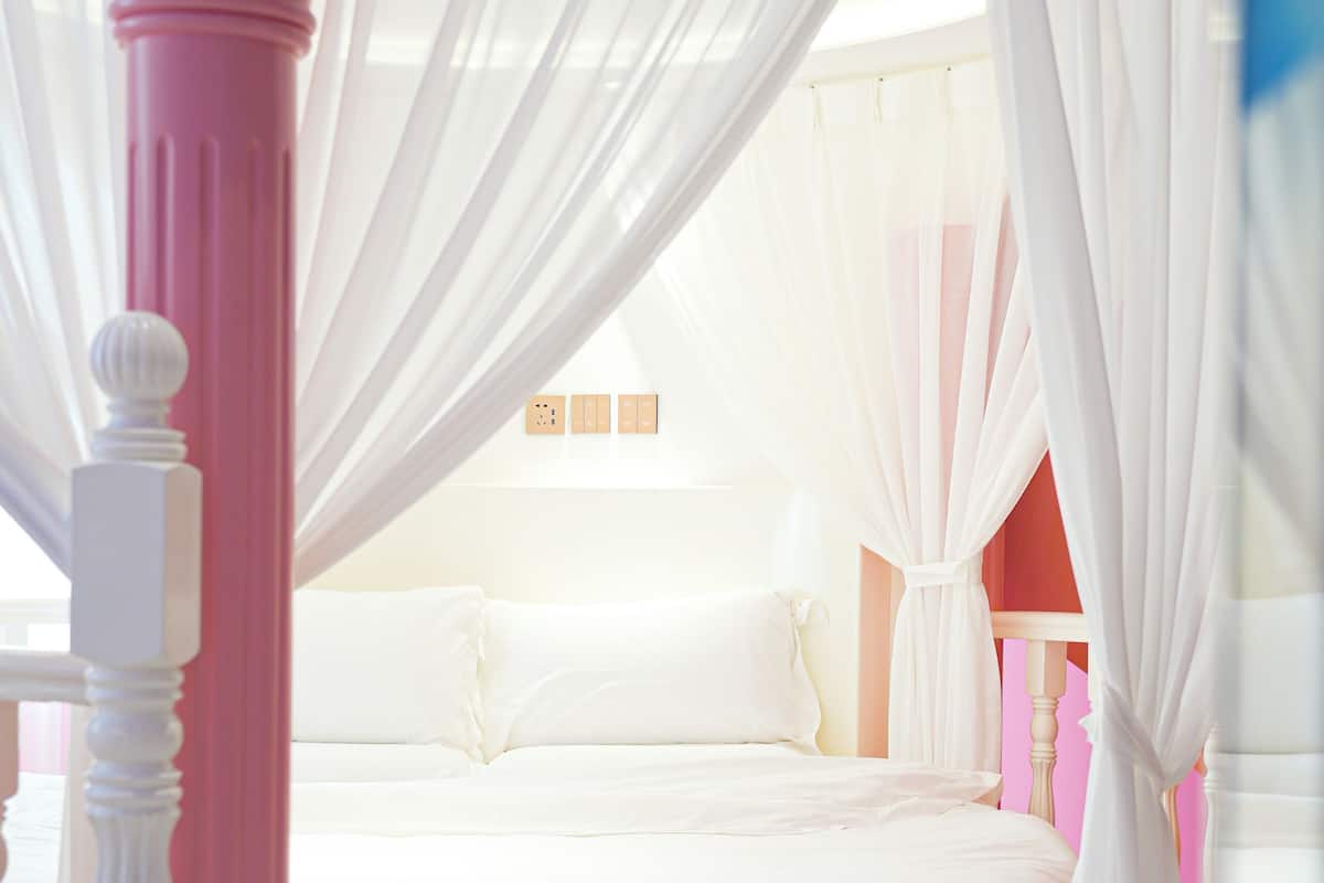 Quarto com cama, cortinas brancas e um pilar rosa.