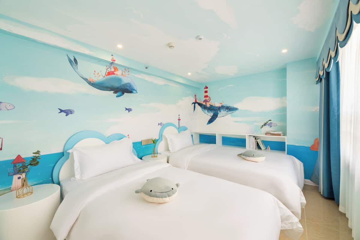 Quarto com duas camas, uma mesa de cabeceira com abajur, e um mural na parede com um farol e criaturas marinhas.
