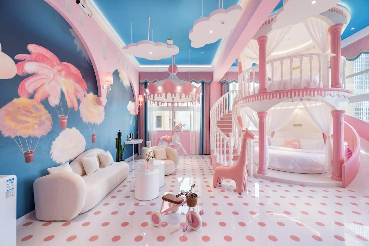 Um quarto com esquema de cores rosa e branco, uma cama redonda com dossel, um sofá branco, uma escultura de girafa rosa e um teto decorado com nuvens.