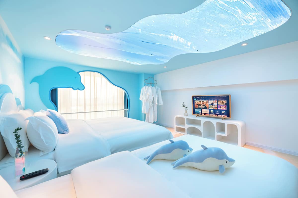 Quarto moderno com uma cama grande, uma cama menor, luminária no teto, televisão e travesseiro em formato de golfinho.