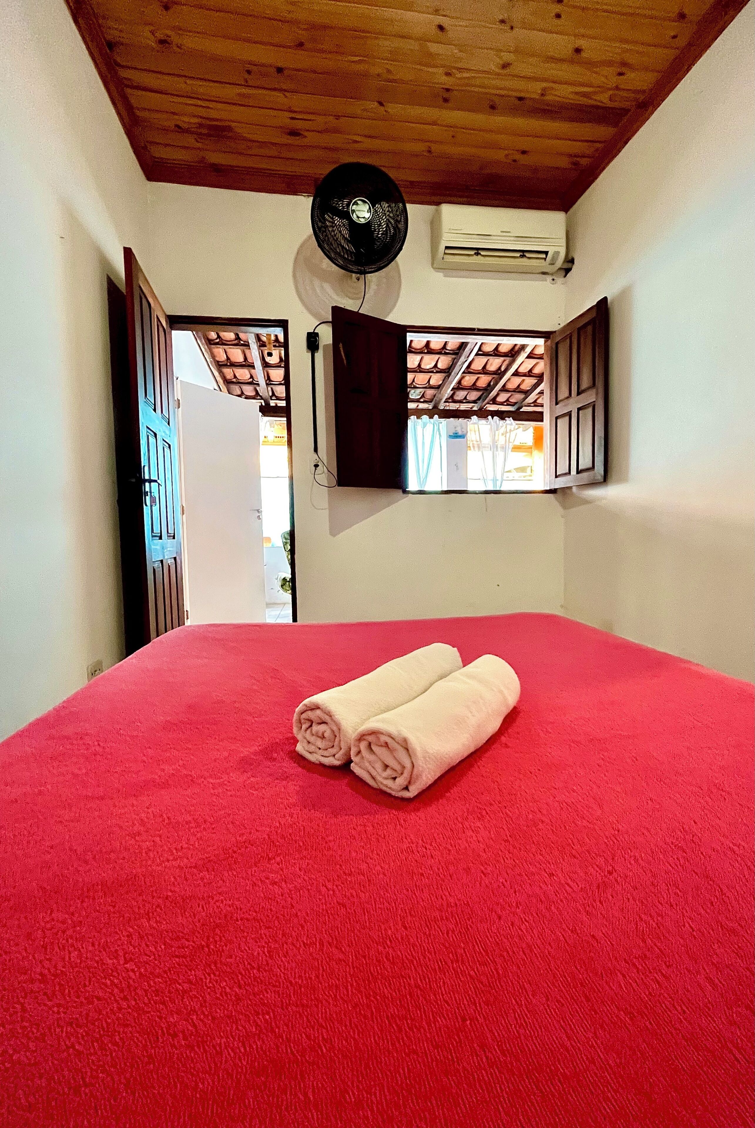 Quarto com colcha vermelha, duas toalhas enroladas, ventoinha no teto, aparelho de ar condicionado e vista através de uma porta aberta.