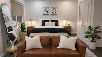 Quarto com cama, duas mesas de cabeceira com luminárias, um sofá de couro marrom, um espelho grande e duas obras de arte penduradas na parede.